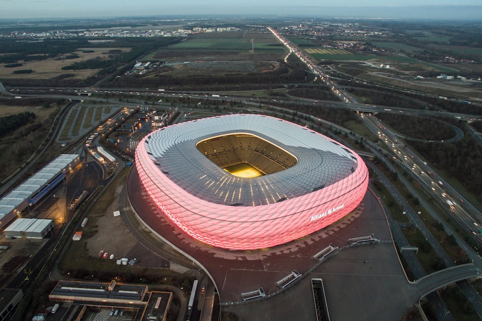 Um estádio grande com um telhado vermelho característico, rodeado por autoestradas e campos.