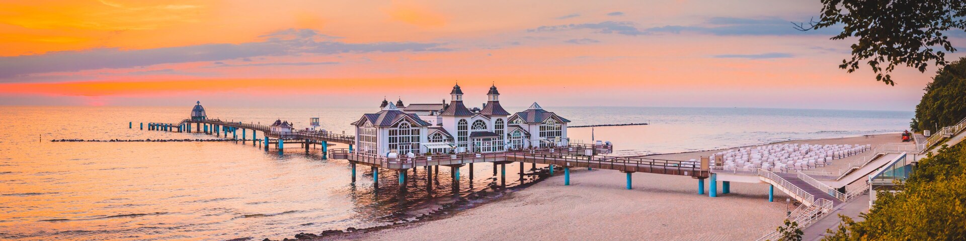 Um cais costeiro com um edifício branco, uma praia e um pôr do sol.