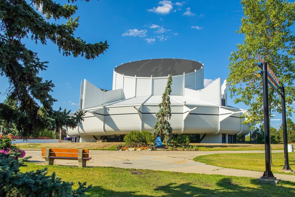 Um edifício moderno com design circular singular, rodeado por um parque bem cuidado com um banco e árvores.