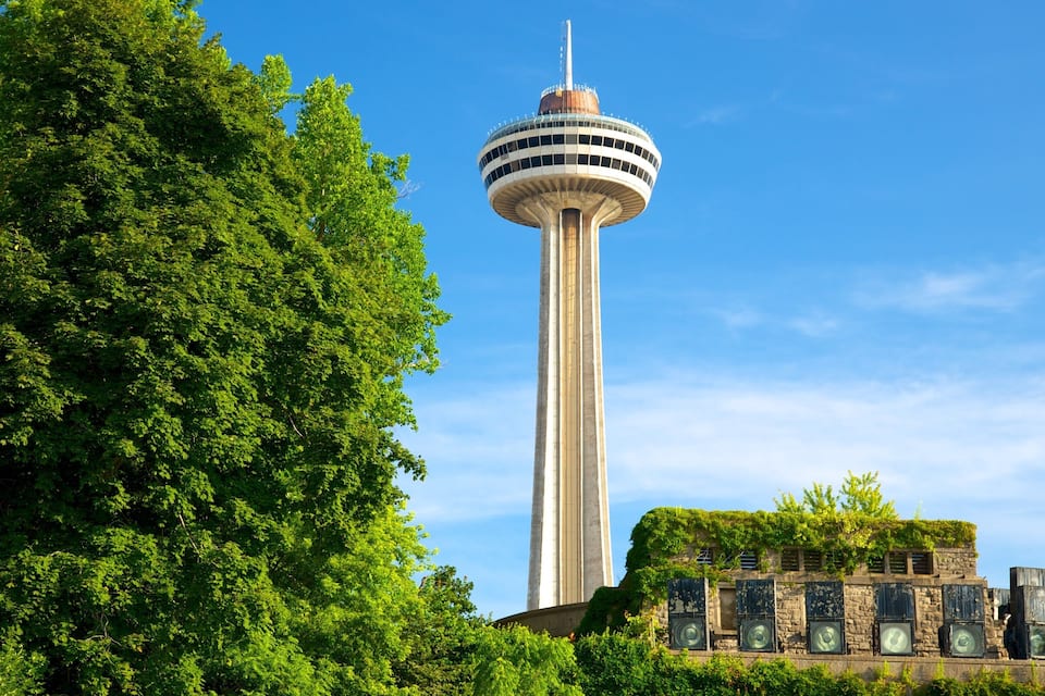 Uma torre com um mirante circular, rodeada por árvores verdes exuberantes e um edifício de pedra com amplas janelas.