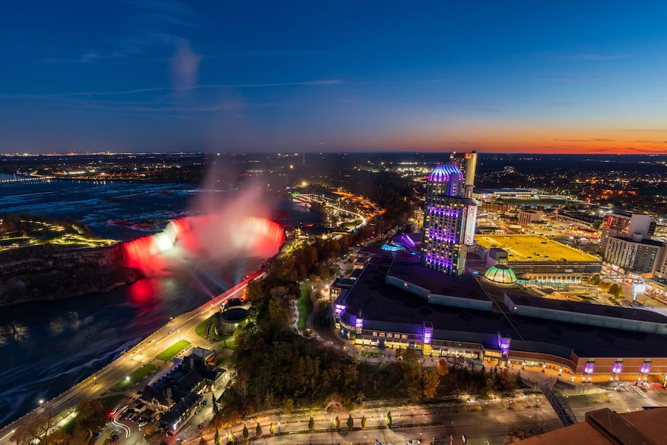 Paisagem urbana ao entardecer com uma cachoeira imponente iluminada em vermelho e branco, um cassino com luzes coloridas e um rio com reflexos da iluminação da cidade na água.