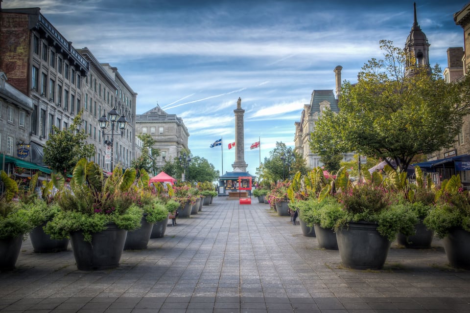 Uma praça pavimentada com plantas em vasos, um monumento e edifícios com bandeiras.