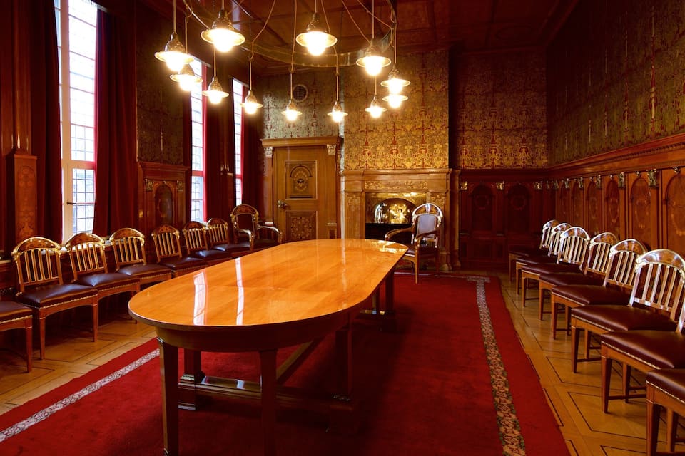 Uma grande mesa de conferência de madeira cercada por cadeiras em um ambiente com papel de parede estampado e lareira.