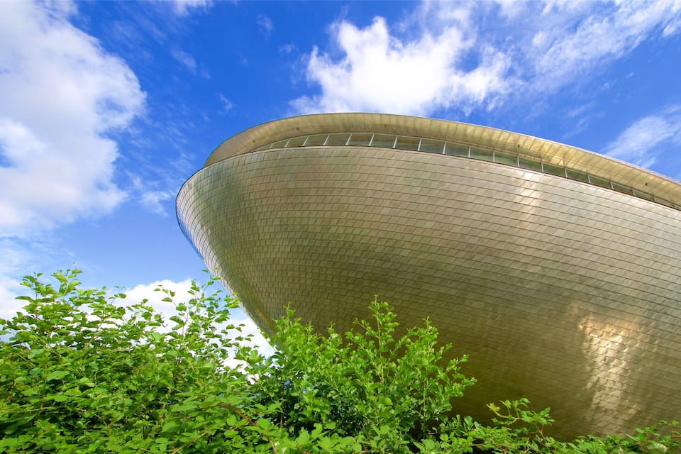 Um edifício grande e curvo com superfície metálica e refletora, rodeado por vegetação exuberante sob um céu parcialmente nublado.