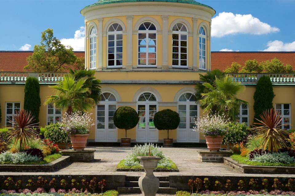 Um edifício amarelo com telhado verde, rodeado por um jardim bem cuidado com diversas plantas e uma fonte central.