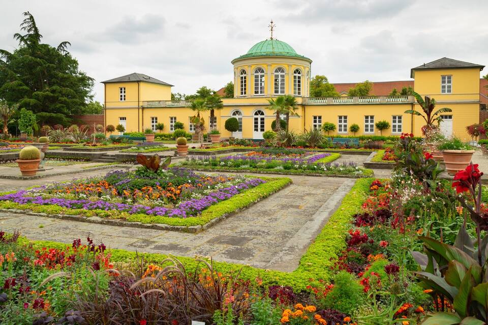 Um edifício amarelo com uma cúpula verde, rodeado por um jardim com várias flores e plantas.