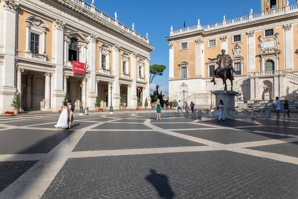 Uma praça histórica com uma estátua de um homem a cavalo, arquitetura clássica e pessoas caminhando.