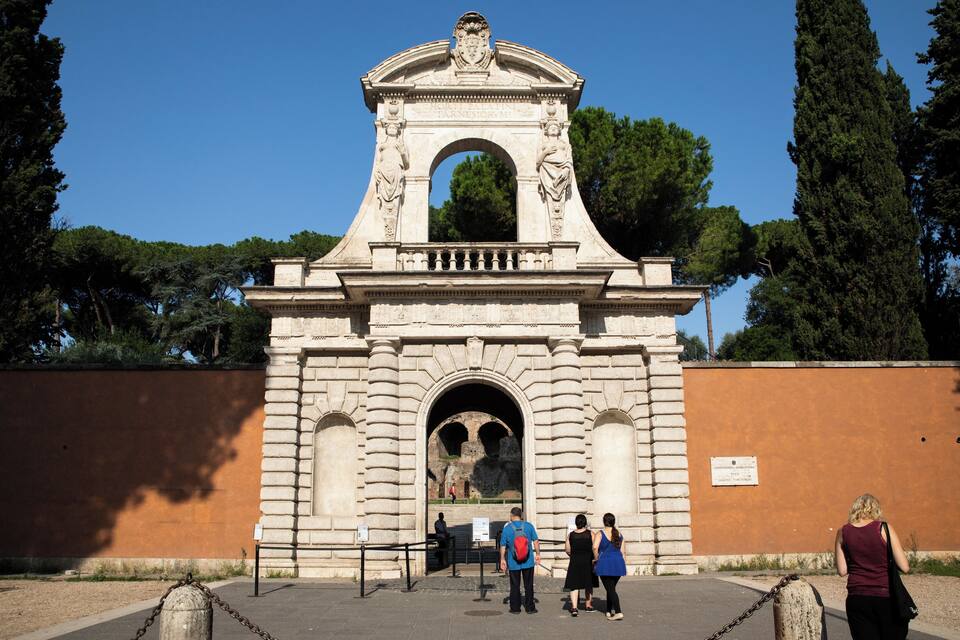 Um arco histórico com inscrições, ladeado por árvores e um muro de tijolos.