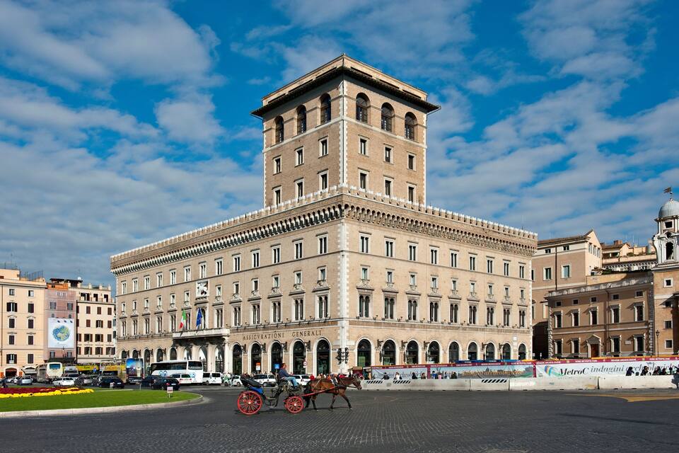 Um edifício histórico com uma torre central e janelas em arco, cercado por uma praça de paralelepípedos e uma carruagem puxada a cavalo.