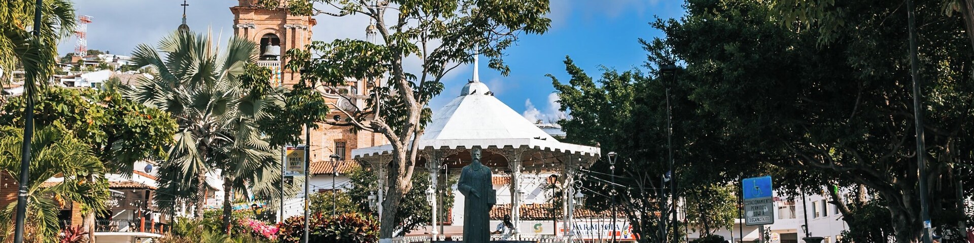 Uma praça com um coreto branco, uma estátua e uma torre com cúpula.