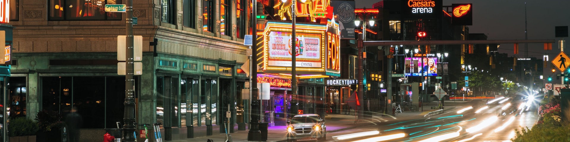 Rua iluminada à noite com letreiros de neon, incluindo um destaque para o letreiro 'Little Caesars Arena'.