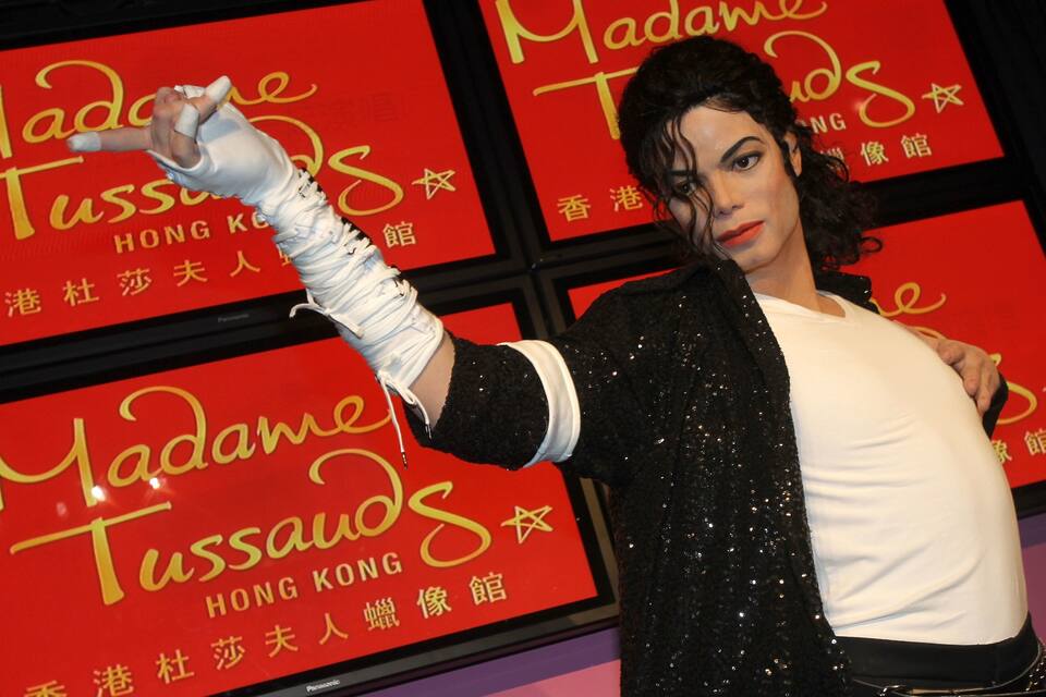 Uma pessoa com casaco preto bordado com lantejoulas e luvas brancas posa diante de um fundo vermelho com a inscrição 'Madame Tussauds Hong Kong'.