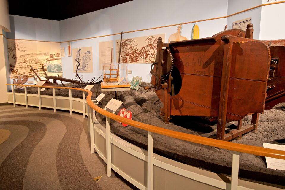 Exposição com artefatos históricos, incluindo uma máquina de madeira grande e diversas ferramentas.
