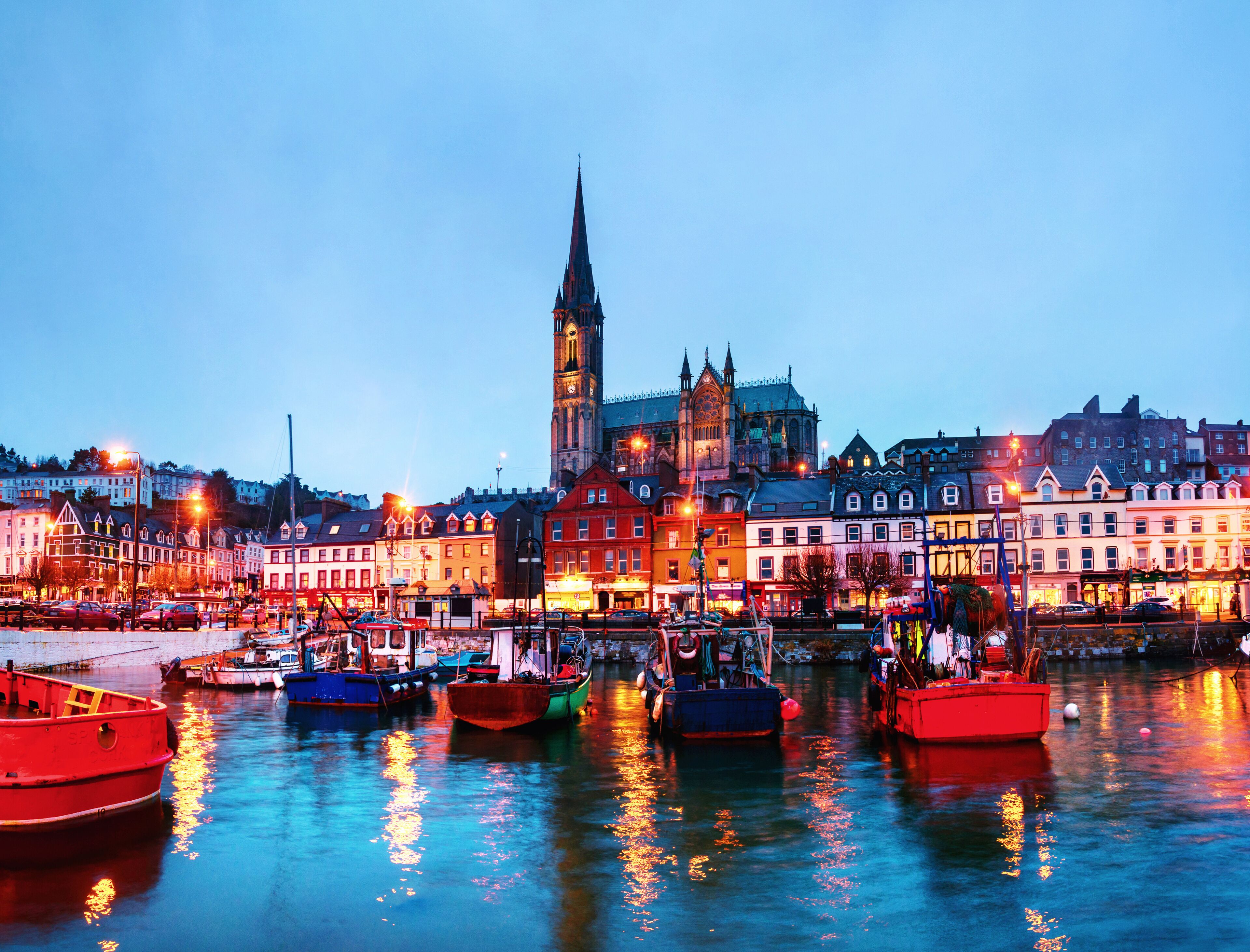 Paisagem urbana à beira-mar com edifícios históricos, uma torre de igreja imponente e barcos atracados refletindo as luzes da noite.