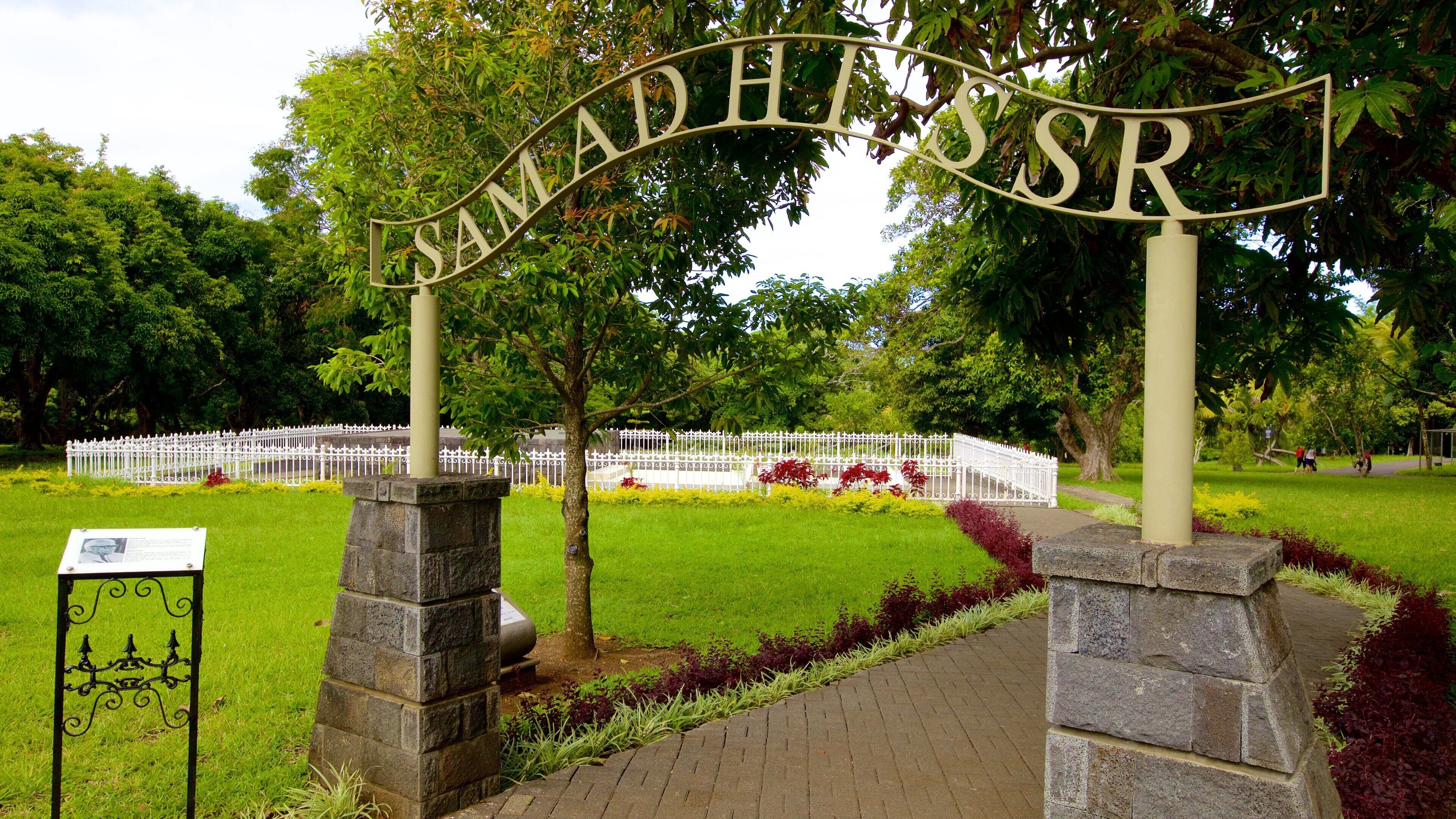 Um arco memorial com a inscrição "SAMAD HESS SR" em um parque com cerca branca, árvores e um caminho pavimentado.