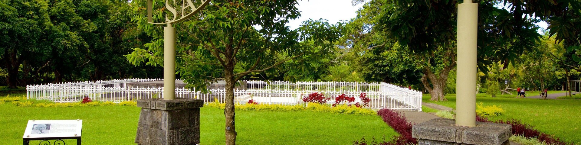 Um arco memorial com a inscrição "SAMAD HESS SR" em um parque com cerca branca, árvores e um caminho pavimentado.