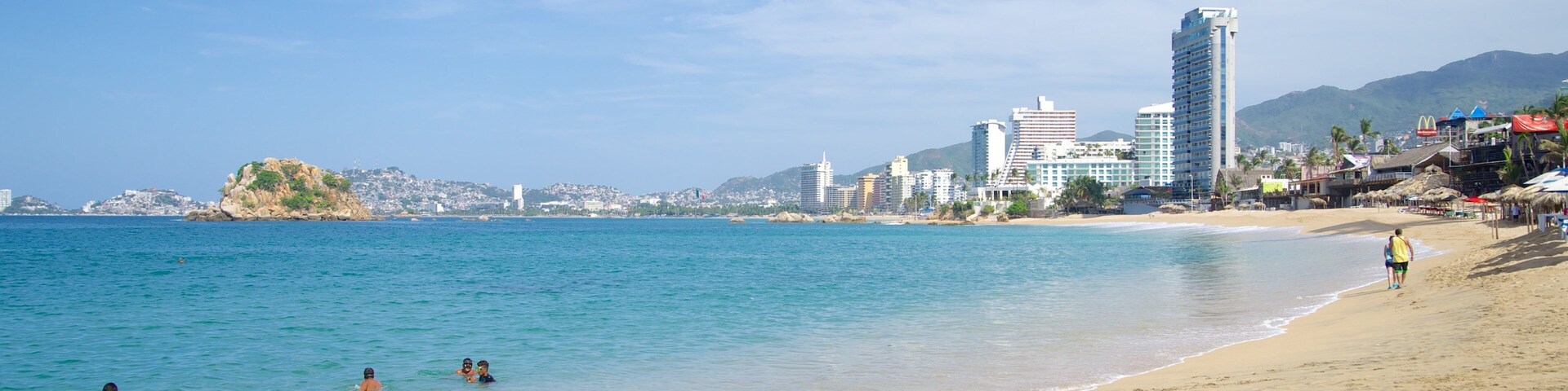 Uma praia com águas azul-claras, areal dourado e um horizonte urbano ao fundo.