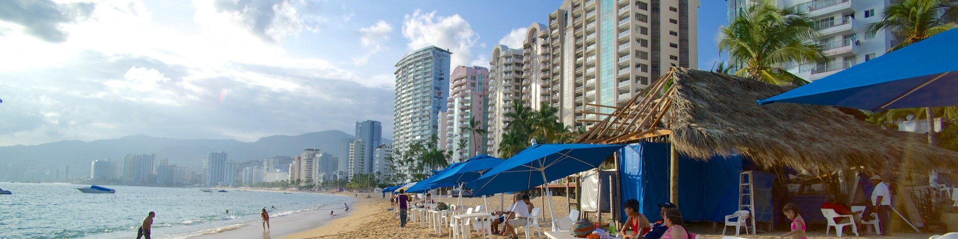 Uma praia com cadeiras e mesas brancas, palmeiras e edifícios altos ao fundo.