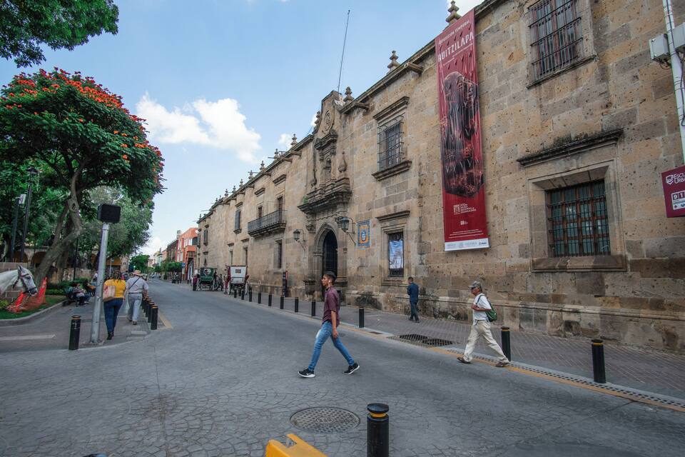 Ruas de paralelepípedos ladeadas por edifícios históricos de pedra, uma árvore com flores vermelhas e pedestres.