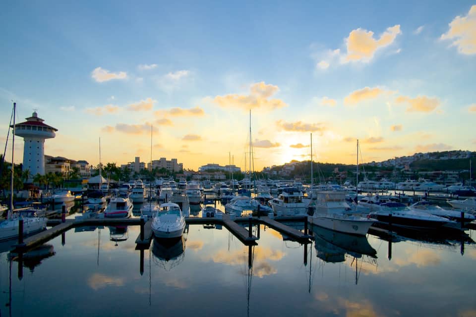 Uma marina com vários barcos atracados, um farol e um pôr do sol ao fundo.