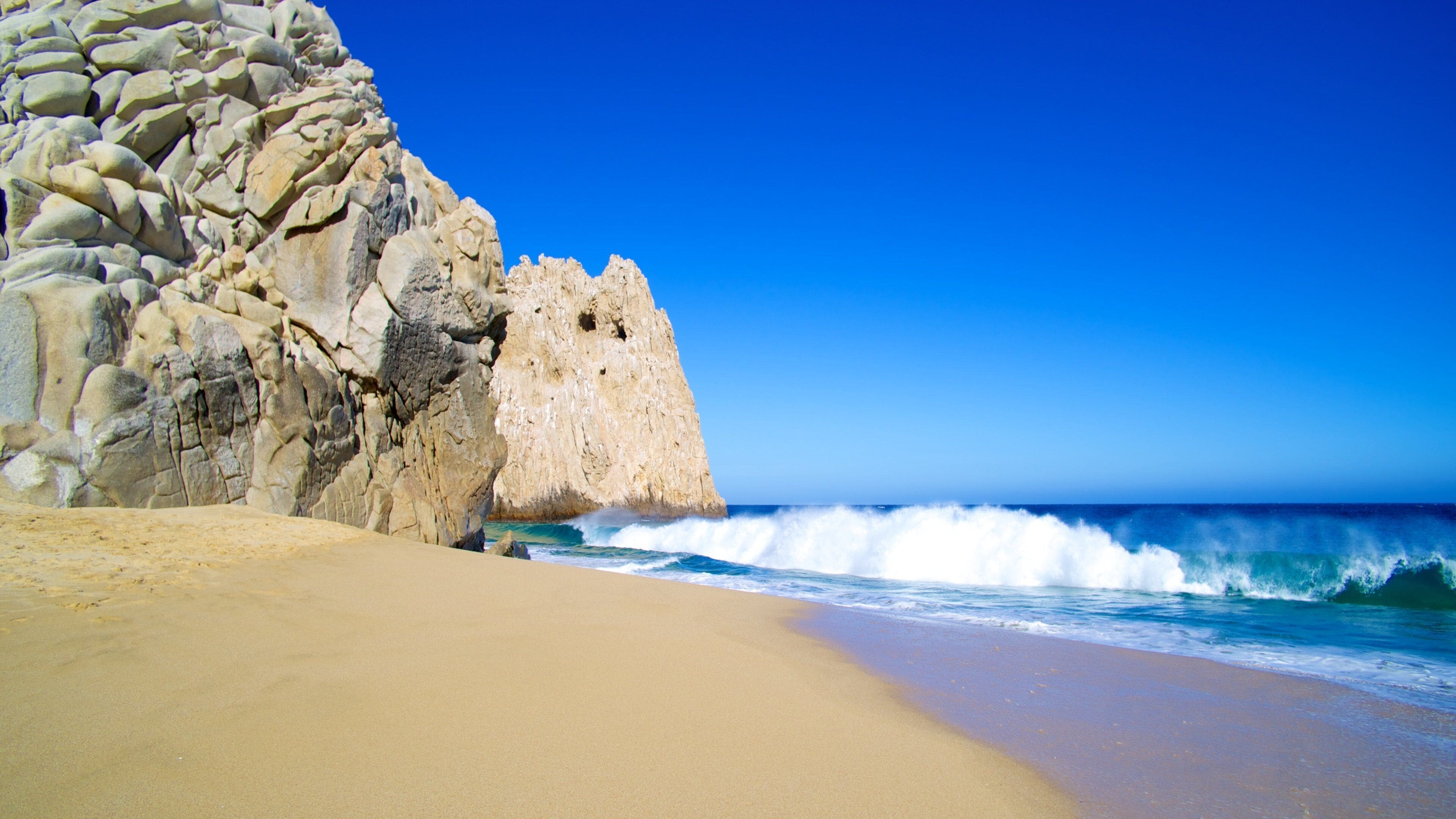 Uma praia arenosa com um penhasco rochoso e ondas quebrando na costa sob um céu azul límpido.