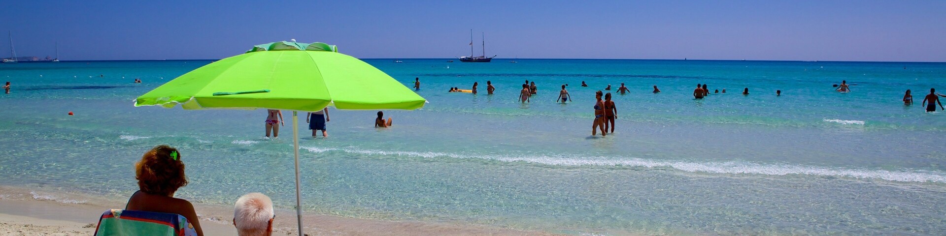 Uma praia com águas turquesa translúcidas, pessoas nadando e caminhando pela orla, e um casal sentado sob um guarda-sol verde.