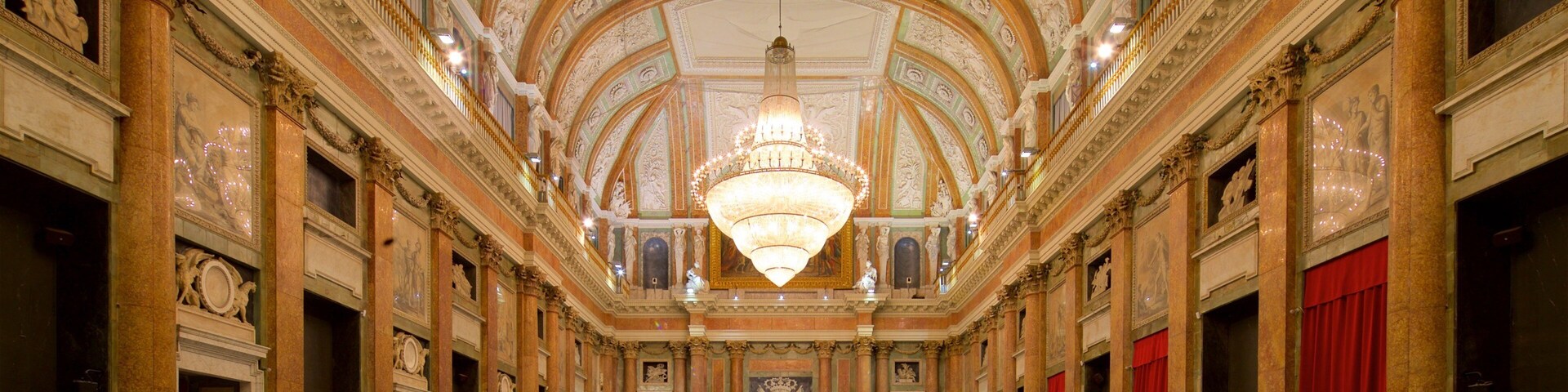 Um salão imponente com colunas decoradas, um grande candelabro e um palco com cortinas vermelhas.
