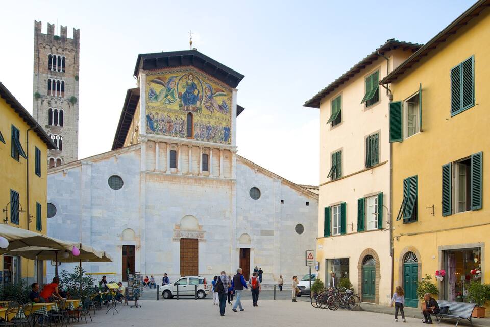 Uma praça histórica com uma igreja destacada, apresentando uma mosaico colorido, rodeada por edifícios tradicionais com persianas verdes.