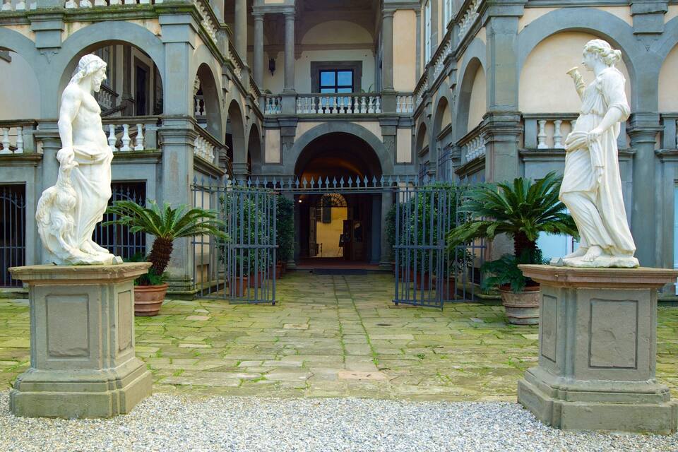 Um pátio com duas estátuas em pedestais, uma entrada com portão e um edifício com arcos e colunas.
