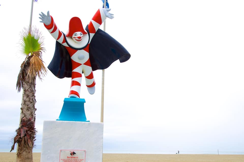 Uma estátua de palhaço sobre um pedestal, com uma palmeira e uma praia ao fundo.