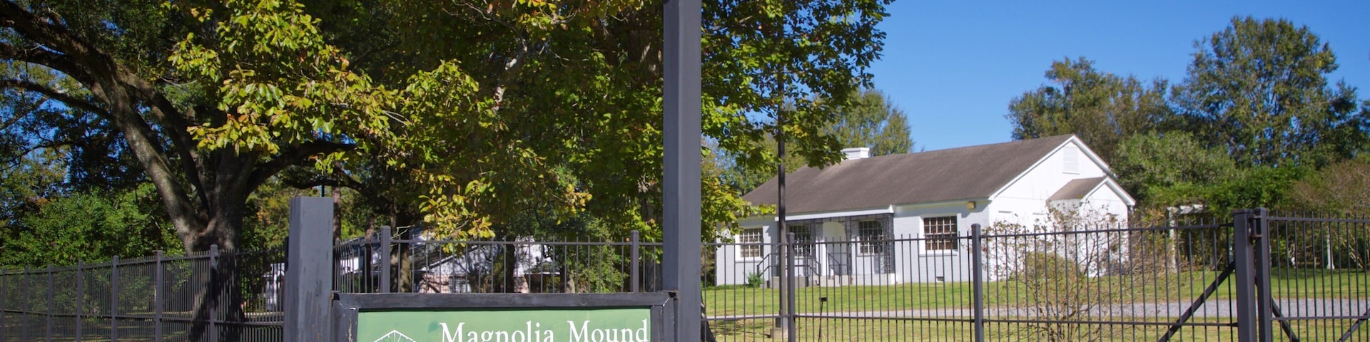 Um sinal verde com letras brancas que diz 'Magnolia Mound Circa 1791' e '2161 Nichols Dr.' em frente a uma casa branca com cerca preta e árvores ao fundo.