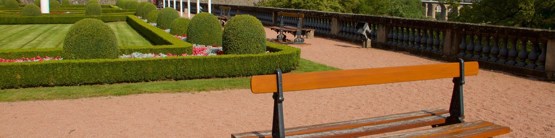 Um parque com um banco de madeira, sebes bem aparadas e um muro de pedra histórico ao fundo.