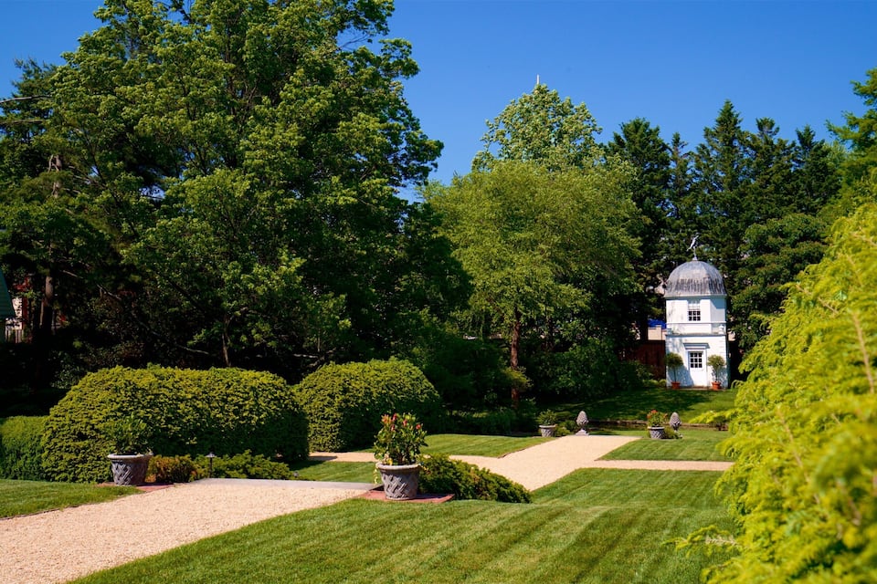 Um jardim bem cuidado com um coreto branco, árvores verdes exuberantes e um céu azul límpido.