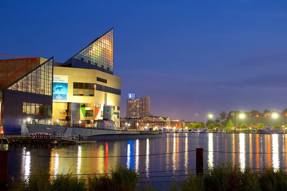Um edifício moderno à beira-mar, com fachada de vidro que reflete luzes, área de cais e horizonte urbano ao fundo.
