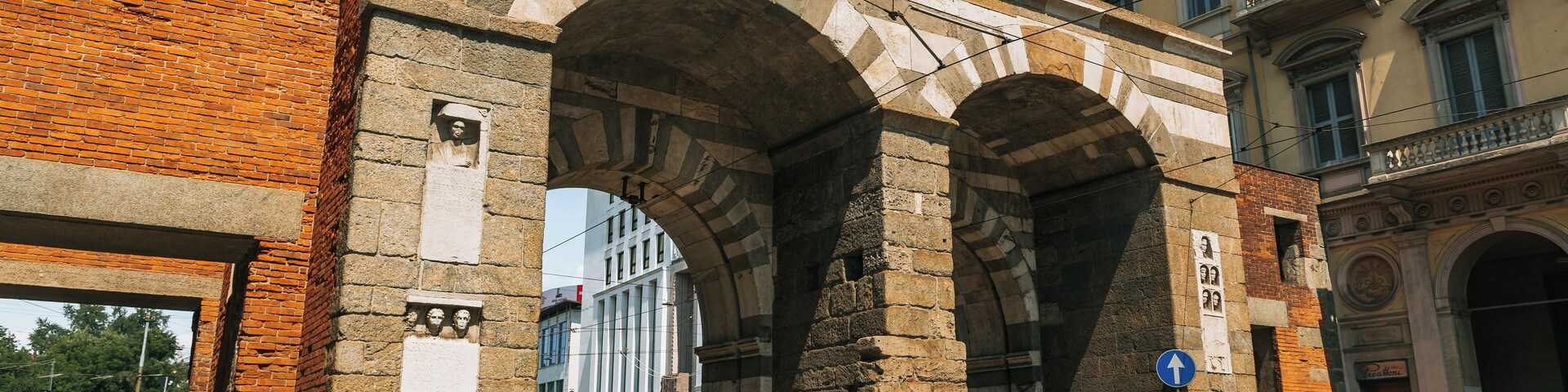 Um arco de pedra com esculturas clássicas, um carro moderno e um edifício com varanda.