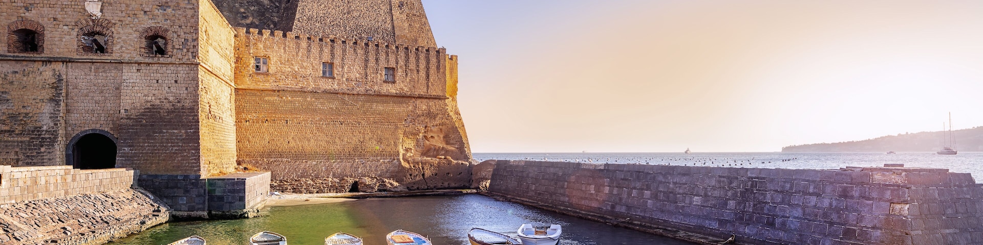 Uma fortaleza de pedra histórica, com fosso, rodeada por barcos coloridos atracados na água.
