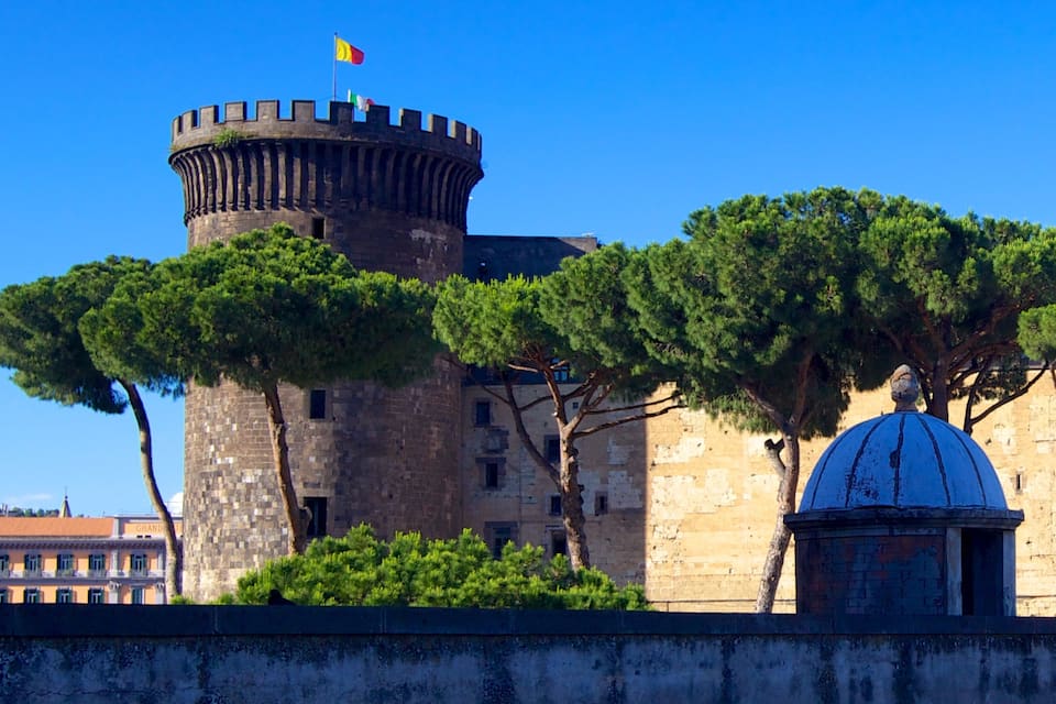 Uma torre histórica com uma bandeira, rodeada por árvores e um edifício com cúpula.