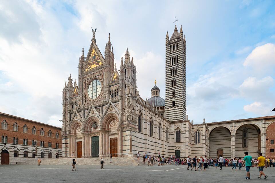 Uma catedral imponente com arquitetura gótica detalhada, uma torre de sino destacada e uma praça movimentada cheia de pessoas.