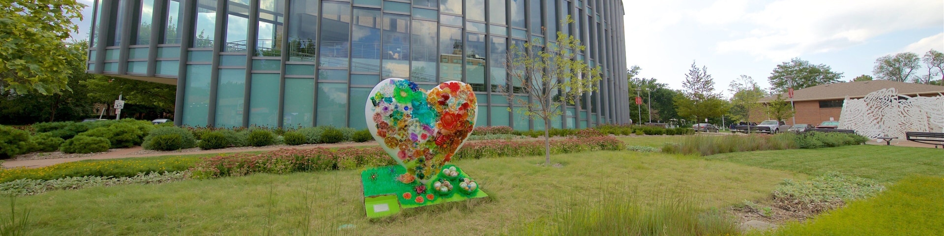 Um edifício moderno com fachada de vidro, uma escultura em forma de coração em uma área ajardinada e uma árvore em primeiro plano.
