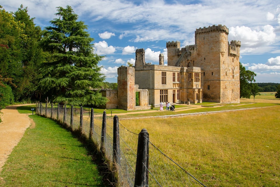 Um castelo histórico com um amplo jardim, um caminho e uma área arborizada.