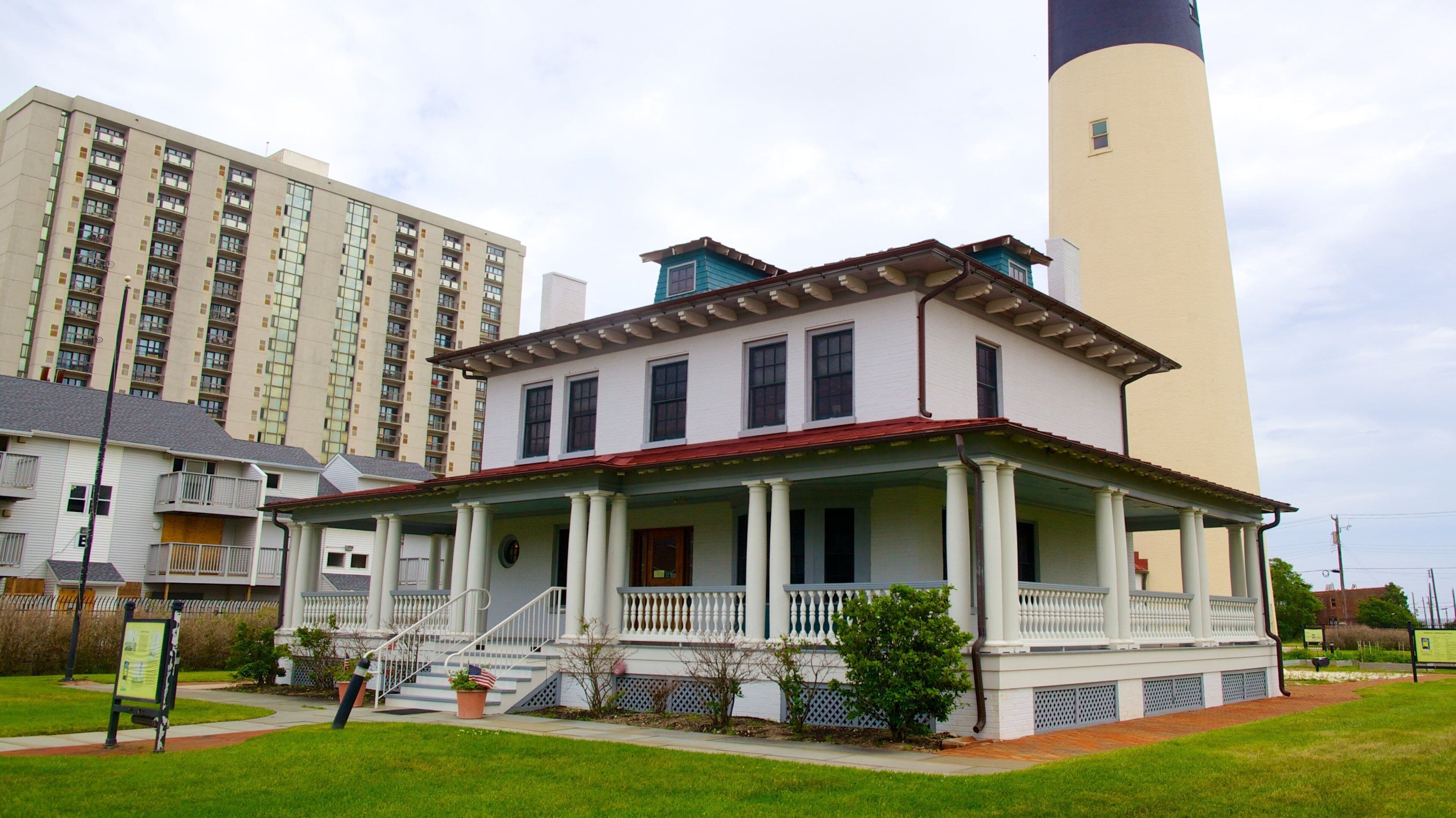 Uma casa histórica com varanda e colunas brancas, um farol alto ao fundo e edifícios de apartamentos modernos por perto.