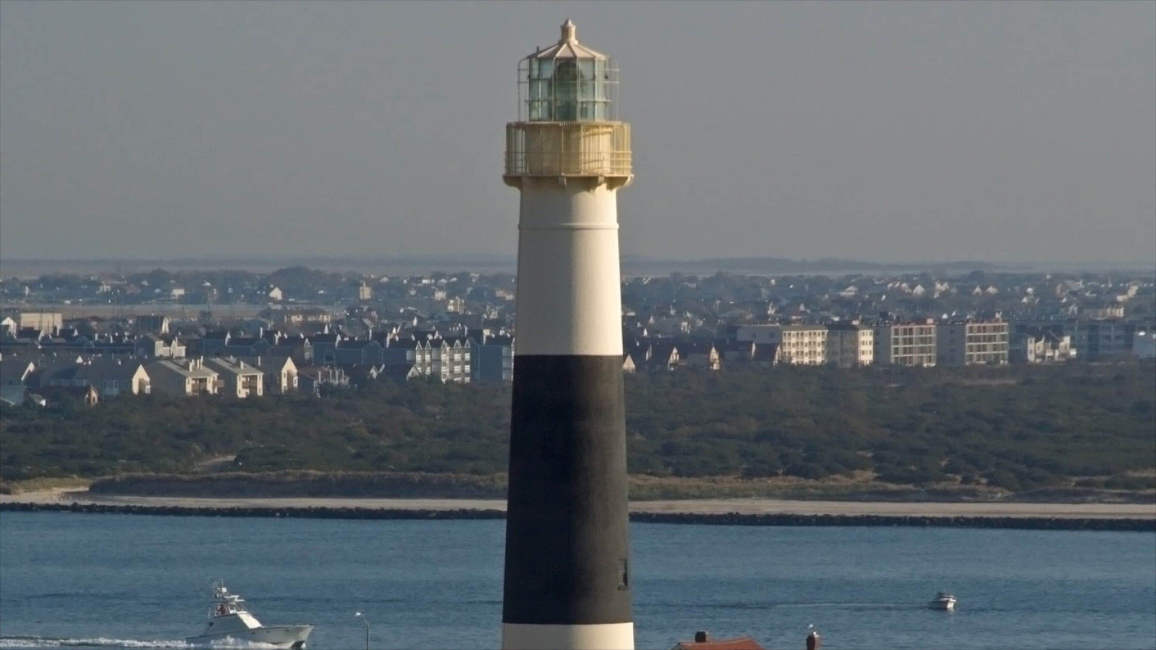 Um farol com torre listrada em preto e branco, rodeado por água e uma paisagem urbana ao longe.