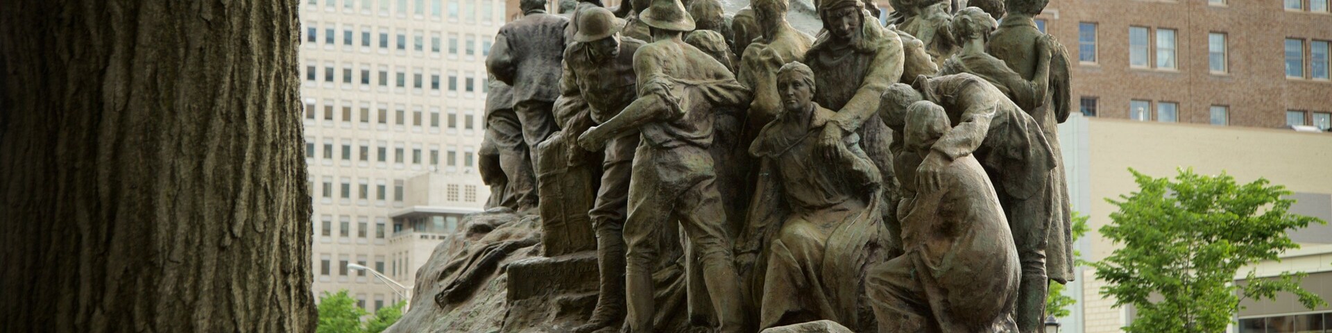 Grande estátua em bronze de um cavaleiro montado com várias figuras, com um cenário urbano ao fundo.