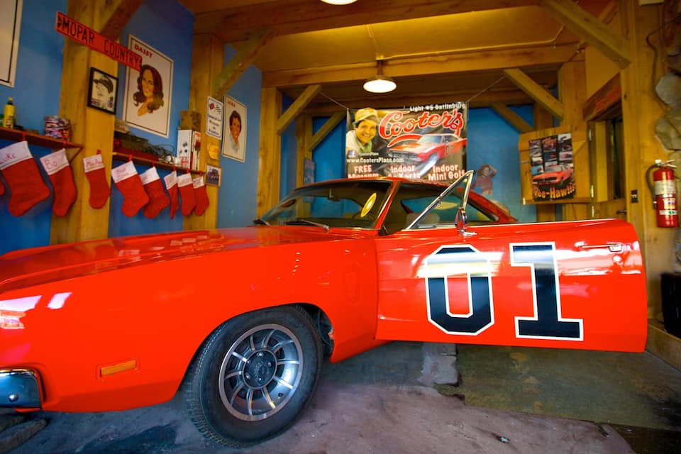 Um carro vintage vermelho com o número 01 em letras brancas, estacionado em um interior com vigas de madeira e diversos cartazes nas paredes.