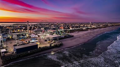 Um parque de diversões à beira-mar ao pôr do sol, com montanha-russa, roda gigante e praia.