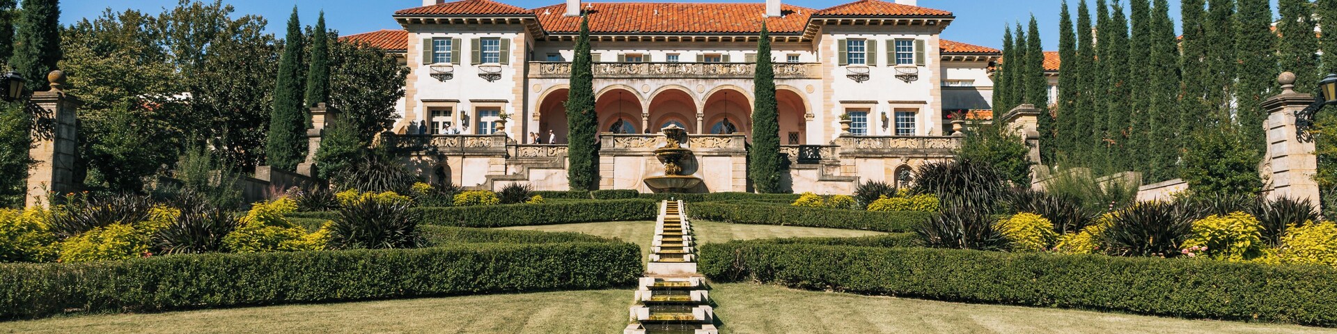 Uma propriedade imponente com um jardim simétrico, uma fonte central e um céu azul límpido.