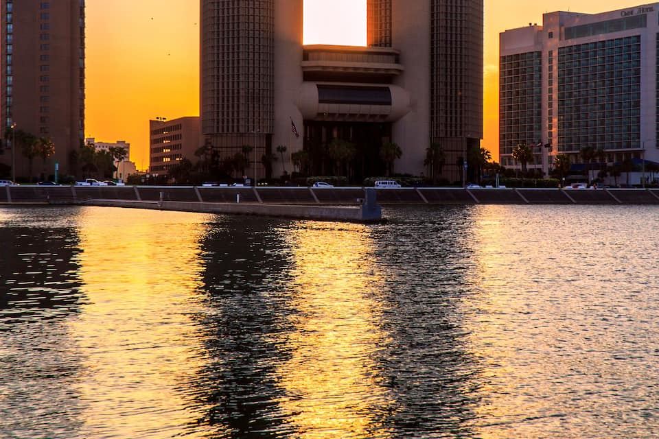 Silhueta urbana com um imponente edifício bancário refletido na água durante o pôr do sol.