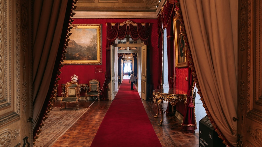 Um corredor imponente com carpete vermelho, mobiliário ornamentado e um quadro grande na parede.