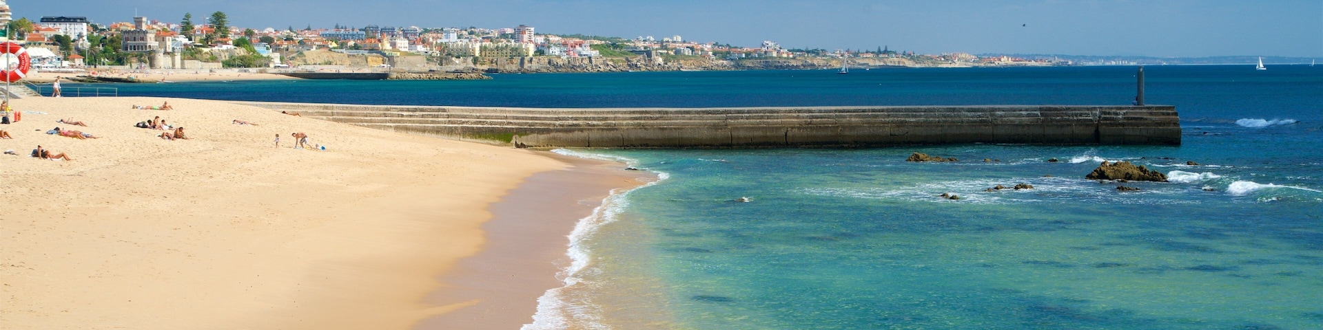 Uma praia arenosa com águas azul-claras, um cais e um horizonte urbano ao longe.