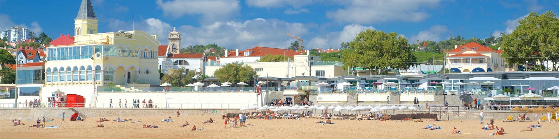 Uma praia com pessoas bronzeando e nadando, uma igreja com torre de sino, e uma cidade costeira com edifícios e guarda-sóis.